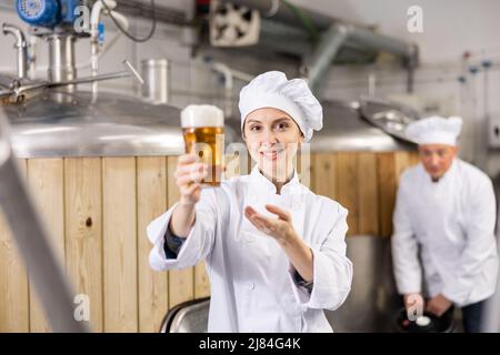 Female brewer is standing with beer with foam in glass on workplace ...
