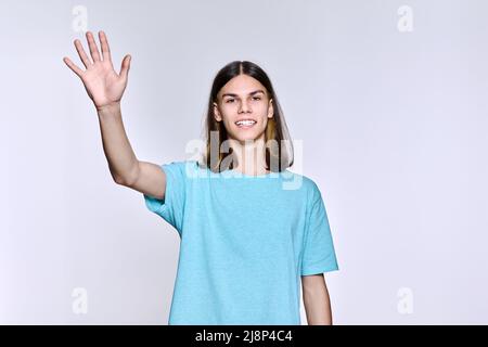 Positive teenage guy waving hand, looking at camera, light studio ...