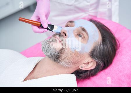 Cosmetologist applying a clay mask to male face with brush. Spa ...