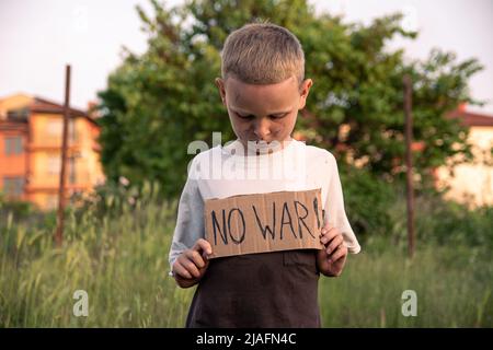 A boy with a grimy face and sad eyes holds a cardboard poster with the ...