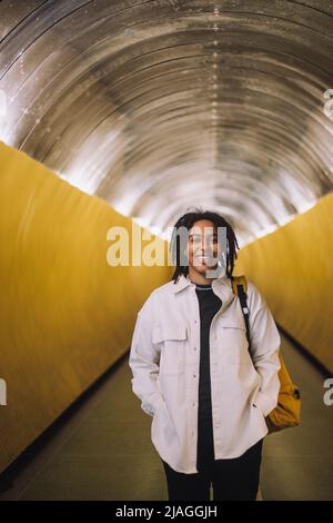 Smiling non-binary person with backpack seen through railings on street ...