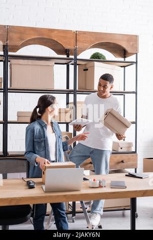 Seller holding scanner near carton box, laptop and colleague in online ...