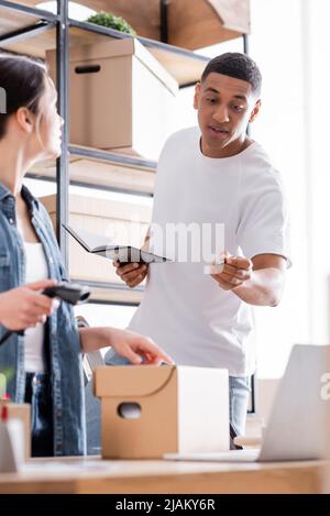 Seller holding scanner near carton box, laptop and colleague in online ...