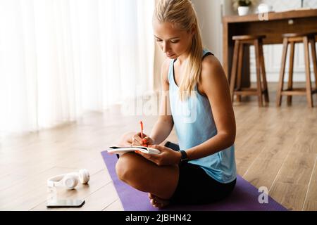 Blonde young woman writing down notes during yoga practice at home ...