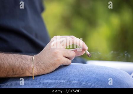 Detail of man holding a lit CBD pre-roll joint, cigarette with smoke ...