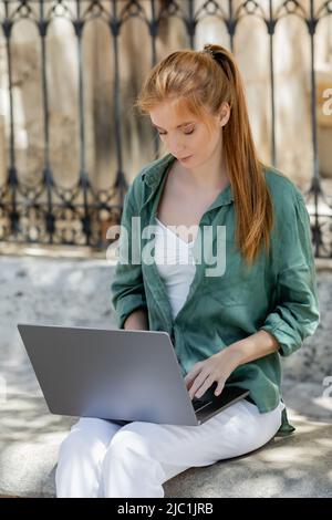 redhead freelancer sitting on concrete bench and using laptop near ...