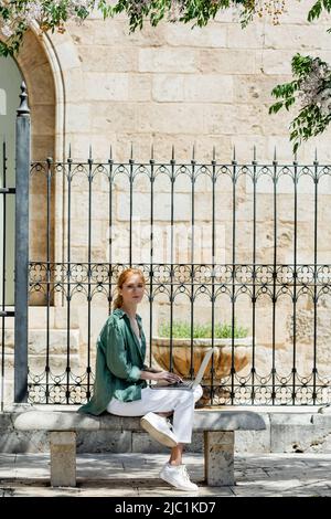 redhead freelancer sitting on concrete bench and using laptop near ...