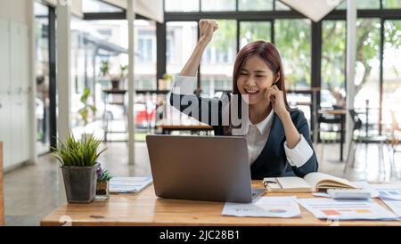 Happy young business asian woman sitting at table with hands up in ...