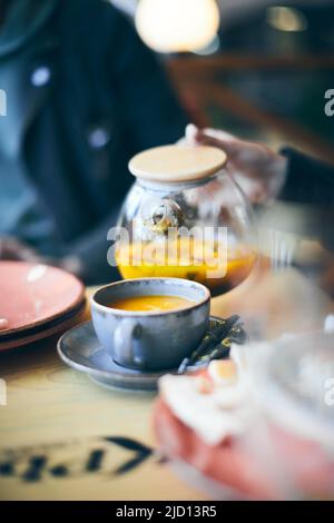 A person pours a yellow liquid from a teapot through a woven strainer ...
