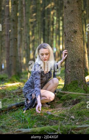Elegant blond woman reaching down to a mossy forest floor as she poses ...