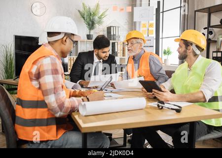Multiracial men builders in formal outfit and helmets sitting at table ...