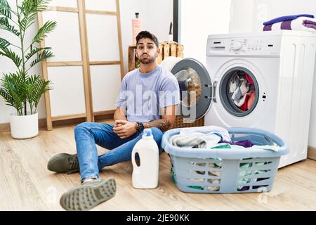 Young hispanic man putting dirty laundry into washing machine clueless ...