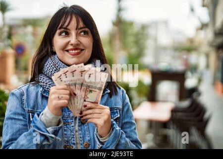 Young hispanic woman holding saudi arabia riyal banknotes scared and ...