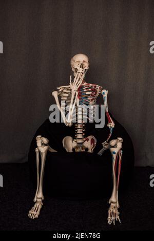 A human skeleton sits on a chair propping up his head with his hand ...