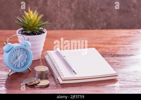 Blank notebook,coins,alarm clock and flower on office desk with wooden ...