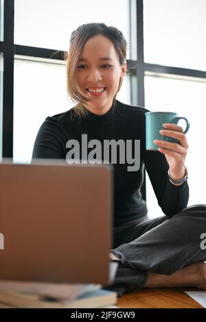 Asian female freelancer smile and holding laptop with rest leg on table ...