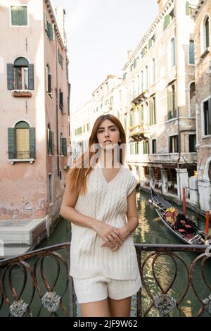 redhead woman in sleeveless jumper looking at camera near colonnade in ...