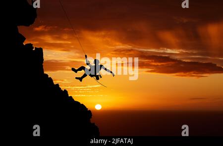 Silhouette of young man abseiling down from a snowy cliff. Climber ...