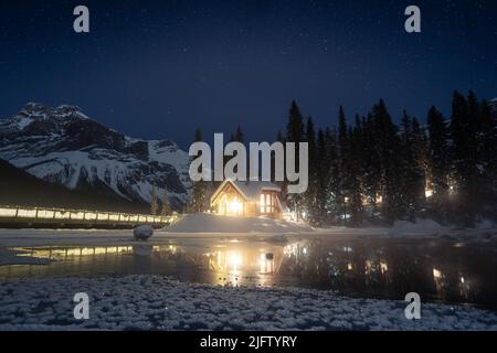 Small hut on the bank of frozen lake surrounded by mountains with stars ...