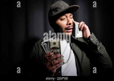 sad indian hip hop performer in cap wiping tear with dollar banknote on ...