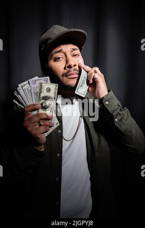 sad indian hip hop performer in cap crying and wiping tear with dollar ...