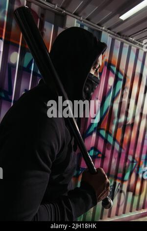 African american vandal in mask holding baseball bat near graffiti on ...