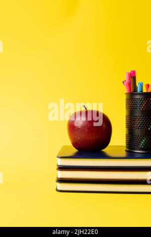 Composition of stack of books, felt tip pens in container and apple on ...