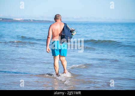 23 July 2022: Rear view of a man walking on the ocean shore with a ...