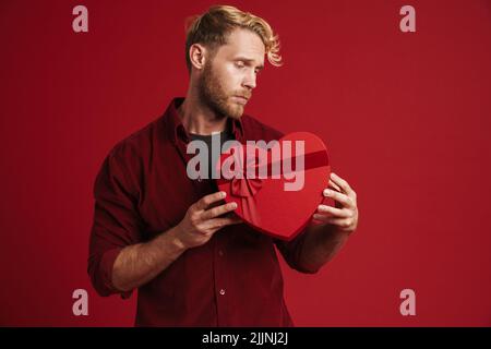 Bearded blonde man frowning while showing heart gift box isolated over ...