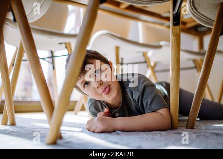 Little boy hiding under the chair at home and making angry grimace ...