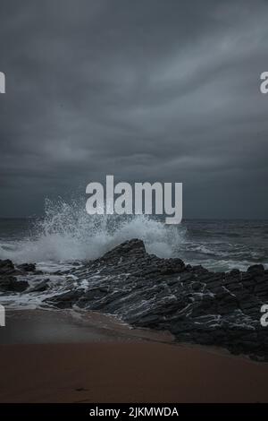 A vertical shot of waves splashing into rocks by the sea during sunset ...