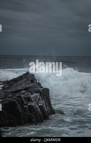 A vertical shot of waves splashing into rocks by the sea during sunset ...