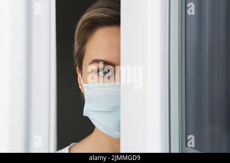 Scared woman wearing prevention mask looking outside through the window ...