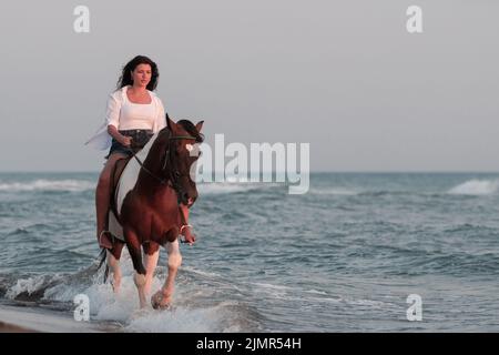 Woman in summer clothes enjoys riding a horse on a beautiful sandy ...