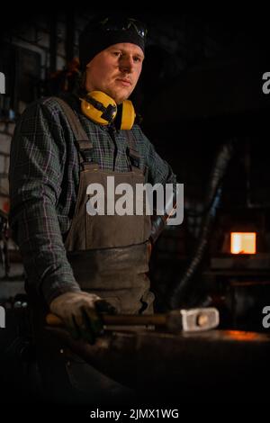 A man blacksmith standing in dark workshop holding a hammer on the ...