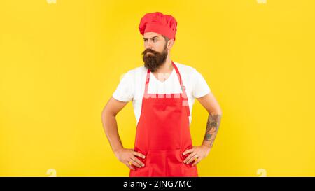 Confident man in red toque and apron looking aside with arms akimbo ...