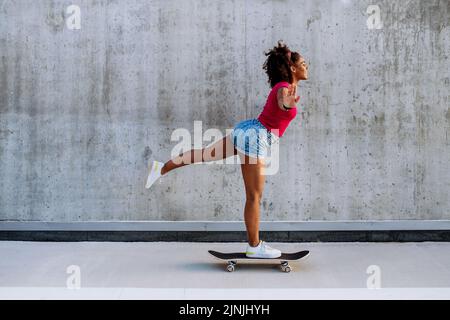 Multiracial teenage girl riding a skateboard in front of concrete wall ...