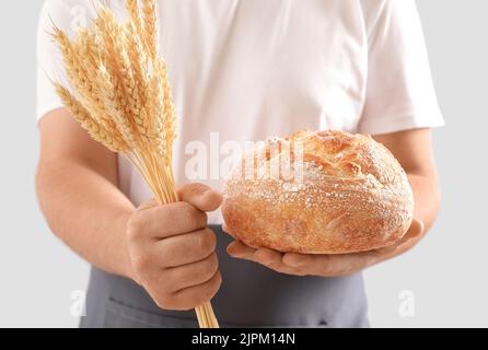 Man with fresh bread and wheat spikelets on light background, closeup ...