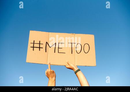 A cardboard sign saying hashtag Me Too held by two hands on clear blue ...