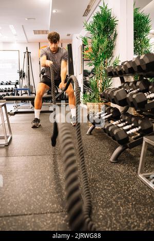Muscular sportsman exercising with rope during sports training in gym ...
