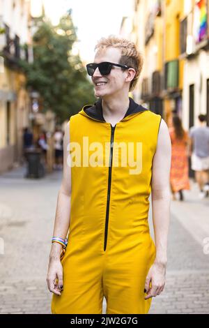 Cheerful transgender young man in yellow jumpsuit looking at camera ...