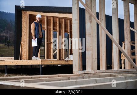 Male inspector building wooden frame house on pile foundation. Man builder standing on ...