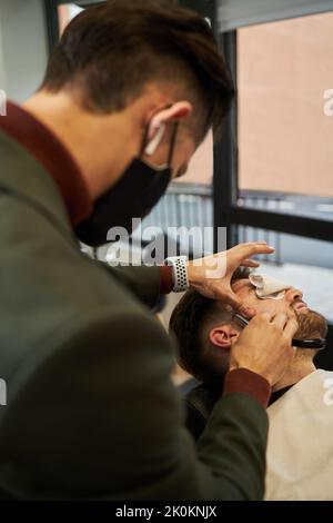 Back view of barber using sharp blade for shaving. Handsome customer ...