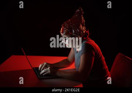 Paranoid caucasian woman typing on a computer wearing a tinfoil cap ...