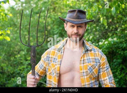 Serious guy holding hayfork. Unshaven guy wearing cowboy hat and plaid ...