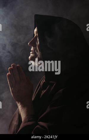 side view of medieval monk in hooded cassock praying on black with ...