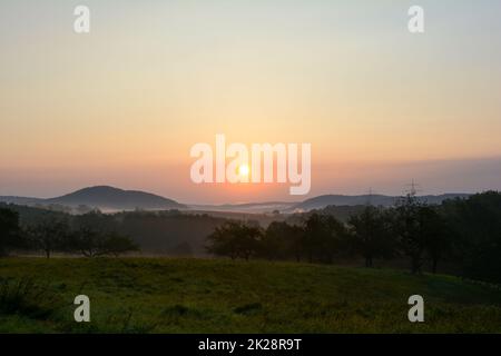 Morning mood at sunrise, hilly landscape with fog, meadow and trees ...
