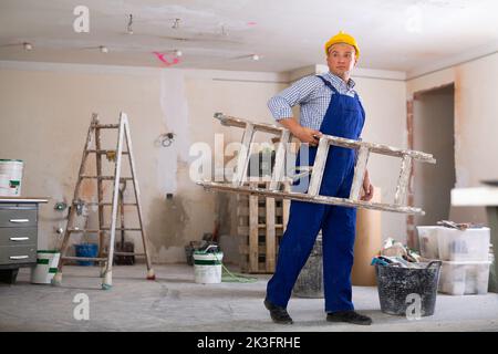 Worker in blue overalls carries a ladder on his shoulder in room being ...