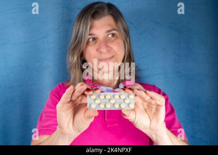A woman holds a pack of pills against a pink shirt and blue background ...