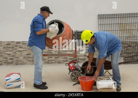 An elderly bricklayer and a young one are mixing concrete with a ...